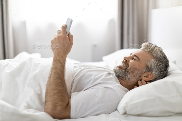 Relaxed happy mature man chilling in bed, using smartphone Stock Photo ...