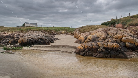 Rocks and beach on ocean coast, farm house on background, dark gray sky ...