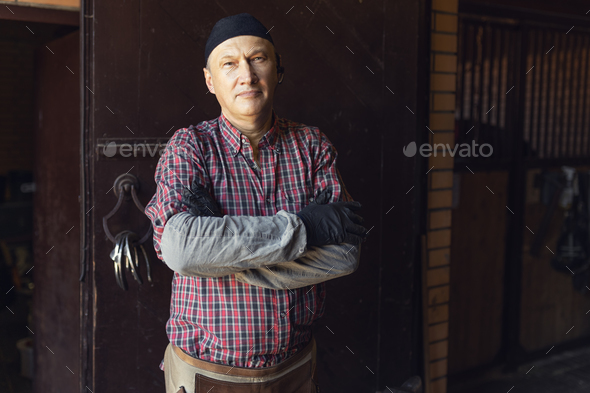 European blacksmith stands at the gate of the stable and his workshop ...