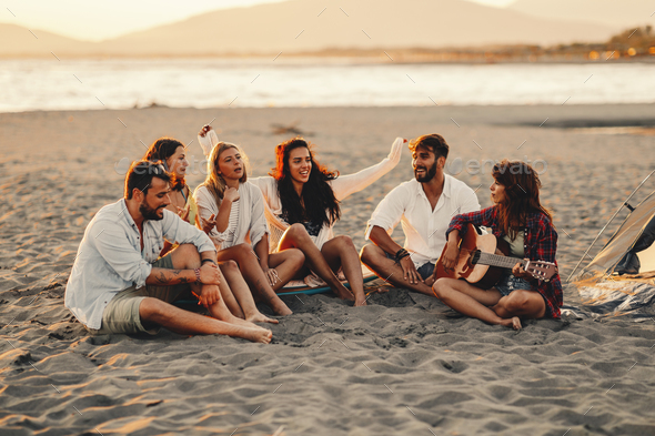 Happy friends sitting on the beach singing and playing guitar during ...