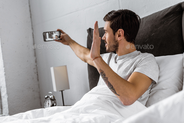 handsome young man chatting with smartphone and waving hand in bed ...