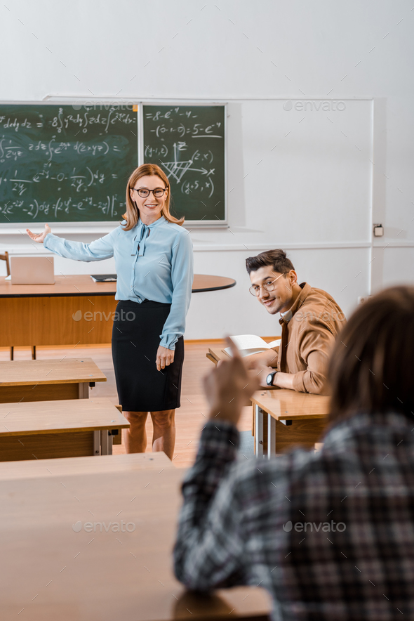 selective focus of smiling female teacher pointing at chalkboard with ...
