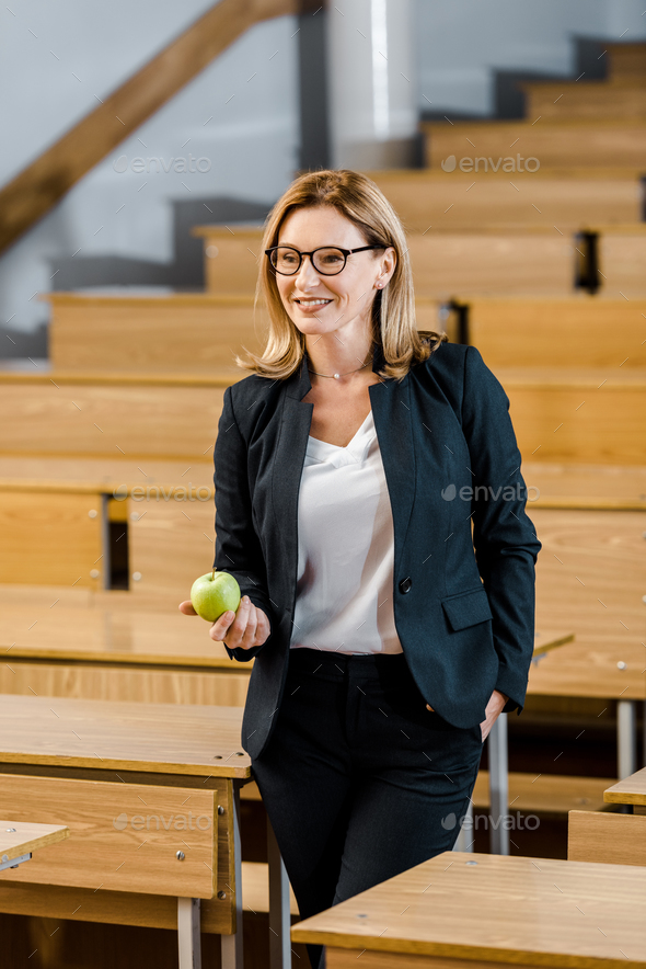smiling female teacher in formal wear holding apple in classroom Stock ...