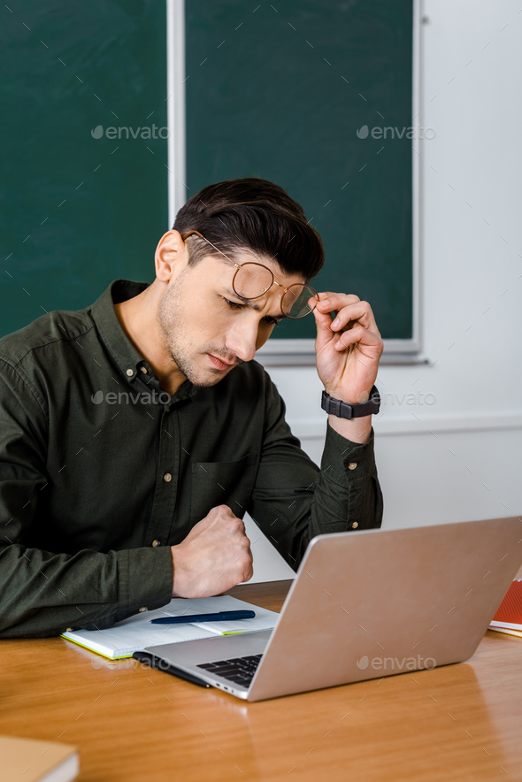concentrated male teacher in glasses using laptop at desk in classroom ...