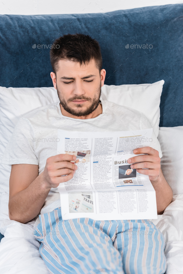 handsome man in pajamas reading newspaper in bed in morning at home ...