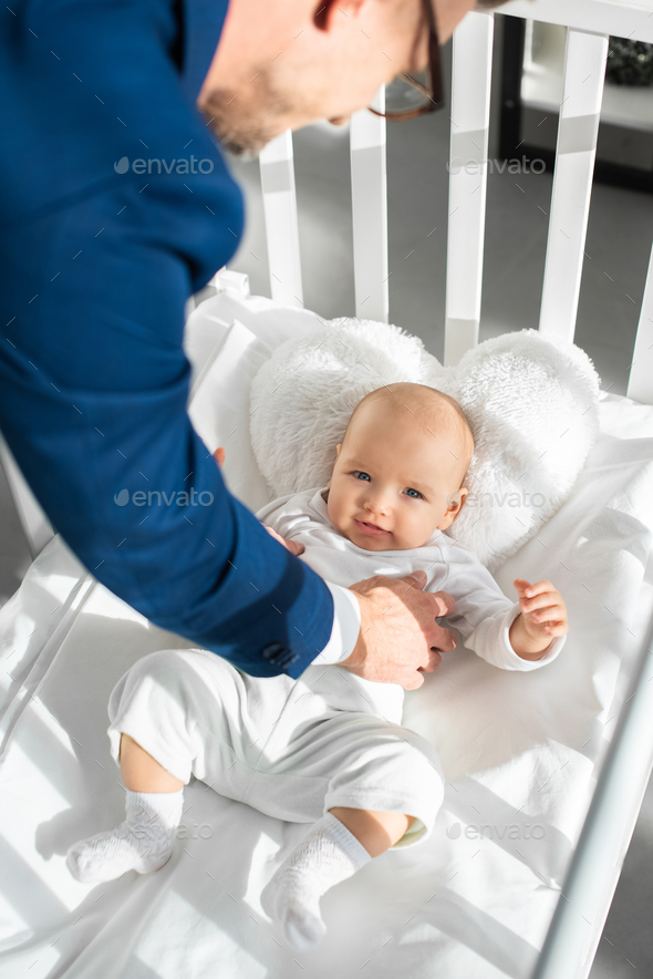 cropped view of father in suit putting infant daughter into baby crib ...