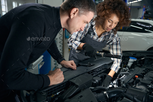 Man in casual clothes and a woman in work overalls look under the hood ...