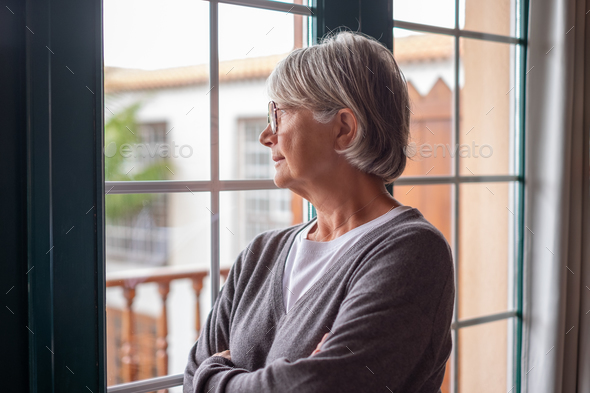Old senior woman standing at window looking outside. Thoughtful old ...