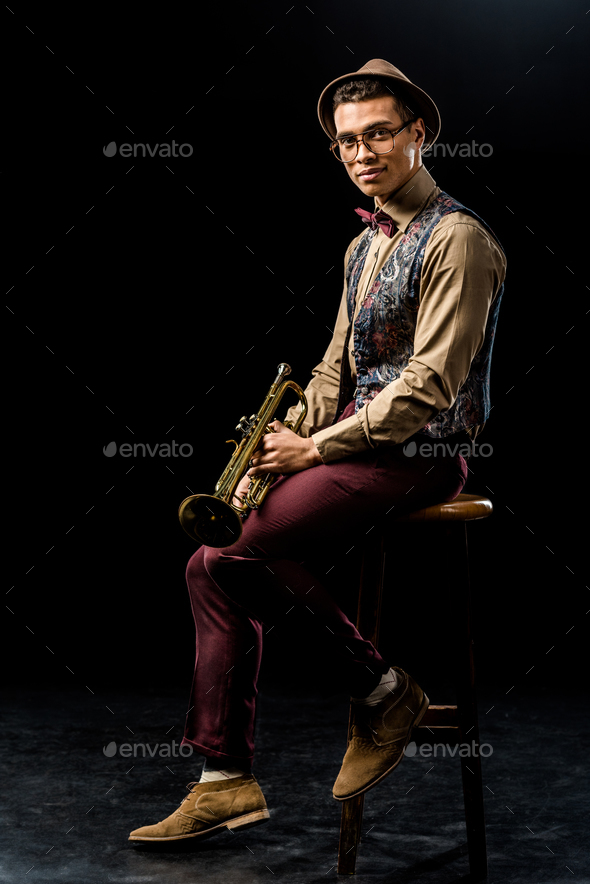 smiling mixed race male musician posing with trumpet while sitting on ...