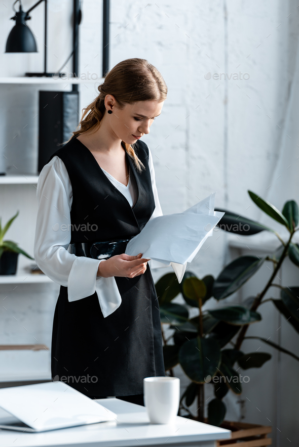 focused businesswoman in formal wear holding documents at workplace ...