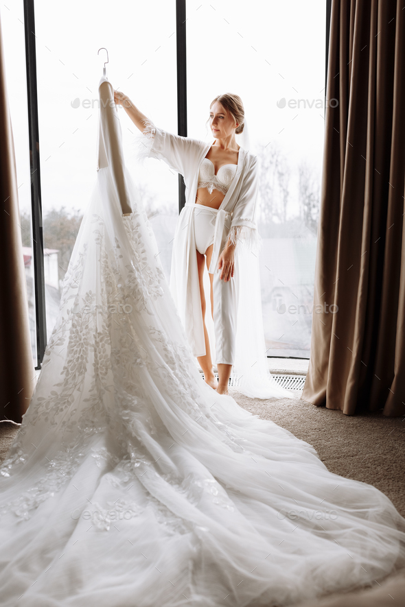 Morning of the bride. beautiful young bride in lingerie is holding a luxury white long dress i ...