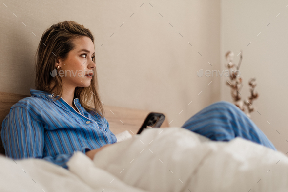 Young woman lying on a bed and scrolling her smartphone. Stock Photo by halfpoint