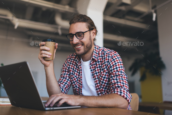 Cheerful programmer in classic eyeglasses watching received video on laptop computer and smiling ...