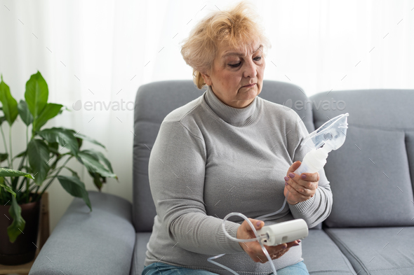 Senior woman using a nebulizer makes inhalation at home and looks at ...