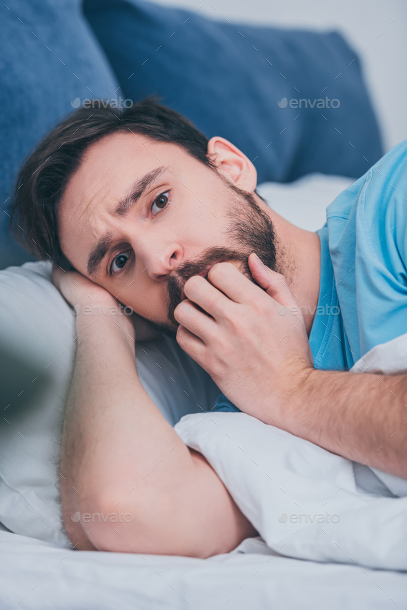 scared man lying in bed, looking at camera and covering mouth with hand ...
