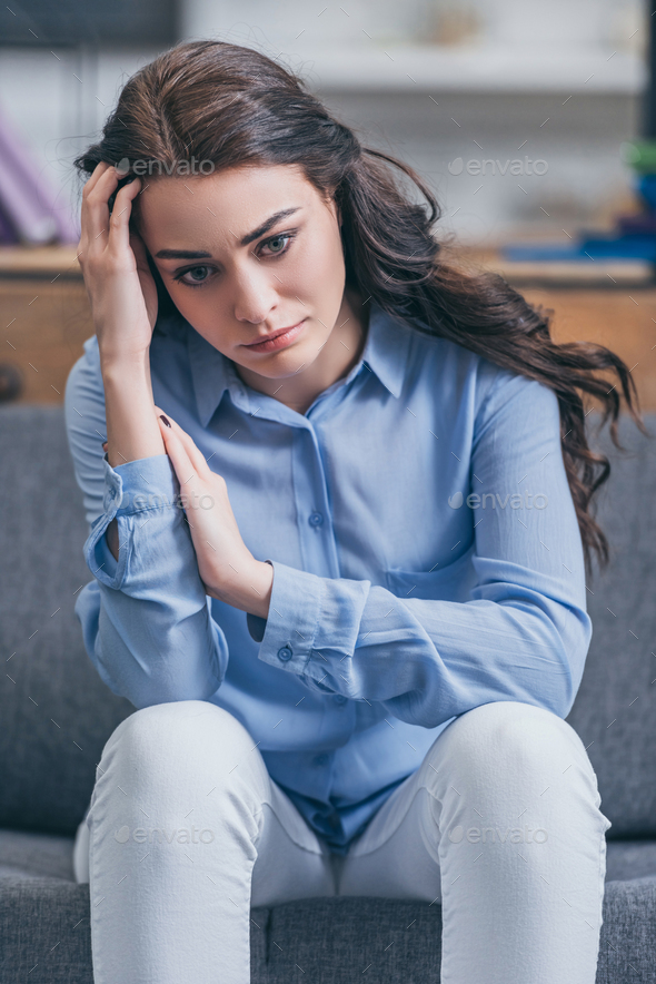 sad woman in blue blouse and white pants sitting on grey couch at home ...