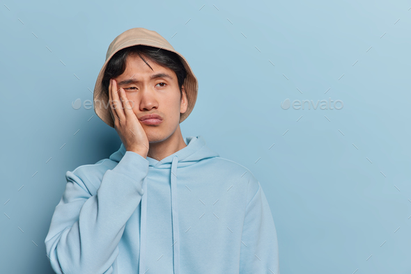 Bored Asian man holds hand on cheek looks tired aside stands in ...