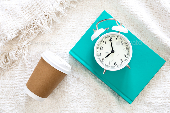 White alarm clock, paper disposable cup and green book in white bed ...