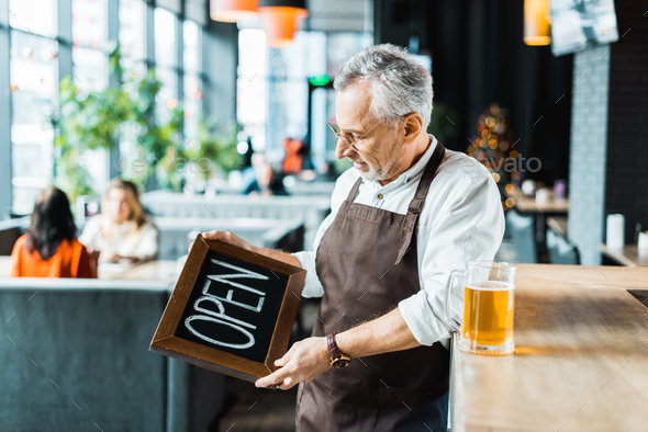 senior owner of pub holding open sign and standing near bar counter ...