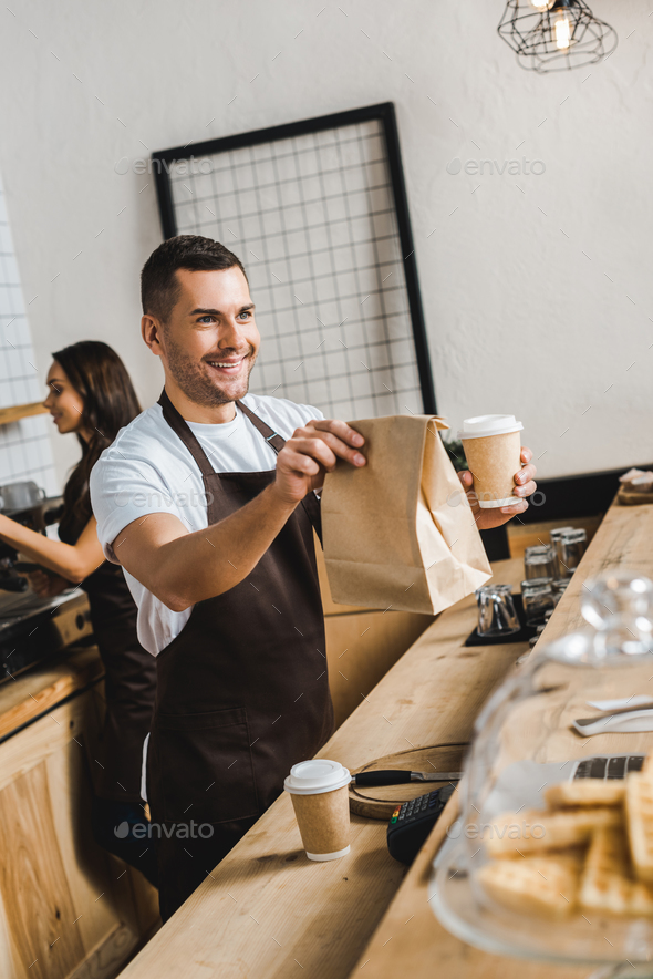 handsome cashier in brown apron giving paper cup and bag wile barista ...