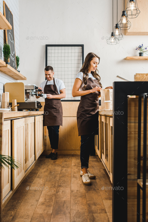 attractive cashier with paper bag and barista in brown apron making ...