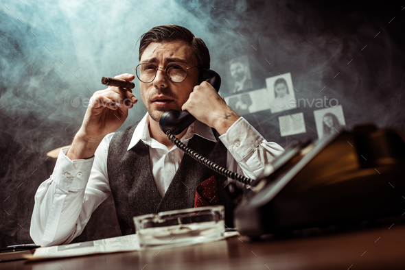 Detective in glasses holding cigar and talking on telephone in dark ...