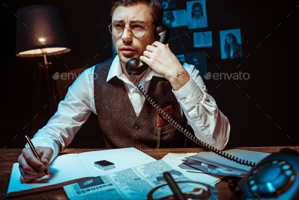 Concentrated detective in glasses talking on telephone and writing in ...