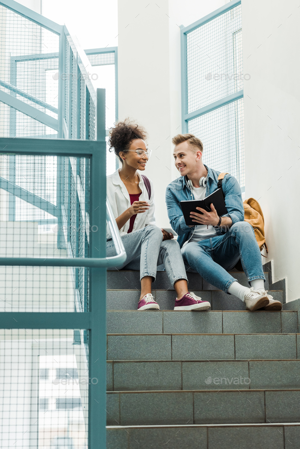 smiling multiethnic students with notebook sitting on stairs in college ...