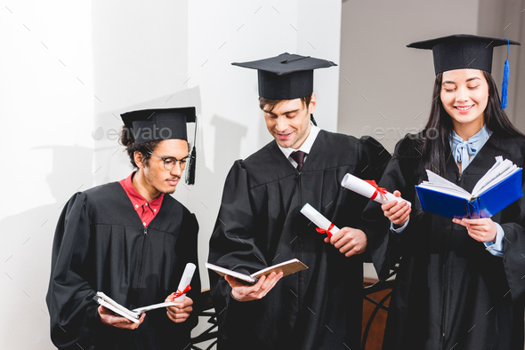happy girl reading book near students in graduation caps holding ...