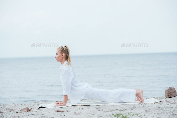 side view of blonde young woman practicing yoga near river on yoga mat ...