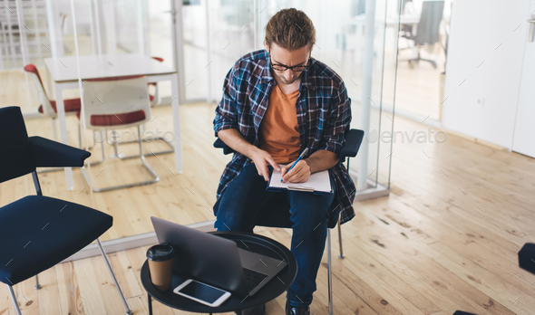 Skilled hipster guy with netbook and paperwork creating design sketches during office work day ...