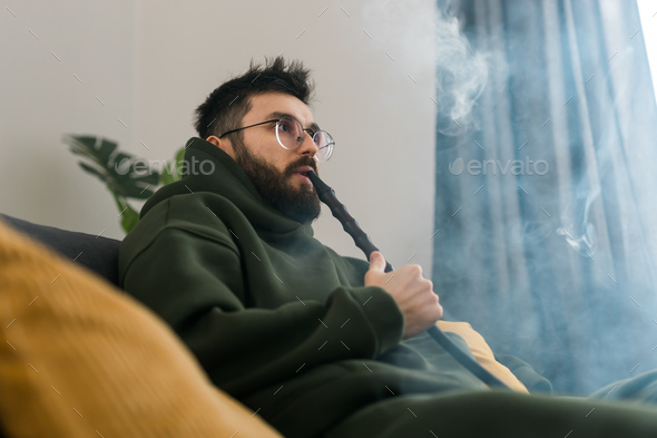 Bearded millennial or gen z man smoking hookah while relaxing on sofa ...