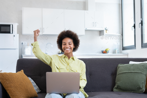 Excited cheerful young Black woman using laptop computer on sofa at ...