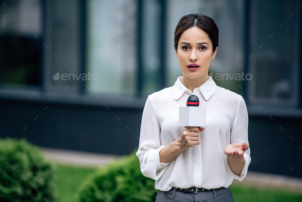attractive journalist in formal wear holding microphone and speaking ...