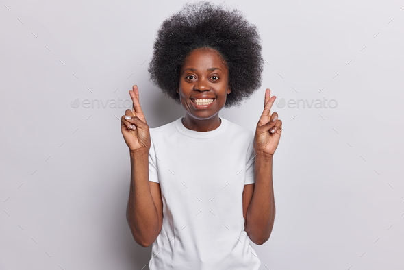 Hopeful dark skinned young woman smiles toothily keeps fingers crossed ...