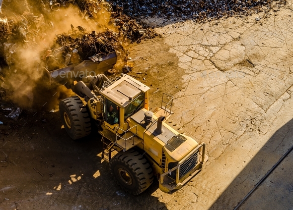 Horizontal shot of a yellow front loader tractor carrying dusty waste ...