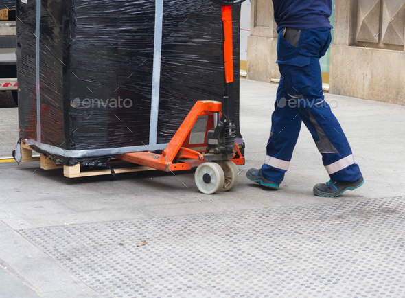 Worker with fork pallet truck stacker in warehouse loading furniture ...