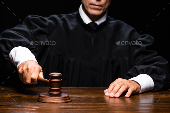 cropped view of judge in judicial robe sitting at table and hitting ...