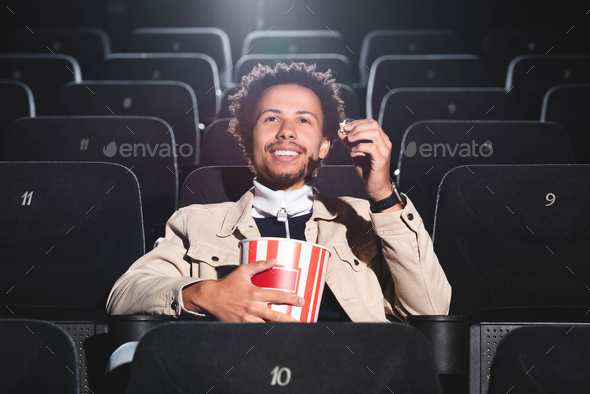 smiling african american man holding popcorn and watching movie in cinema Stock Photo by ...