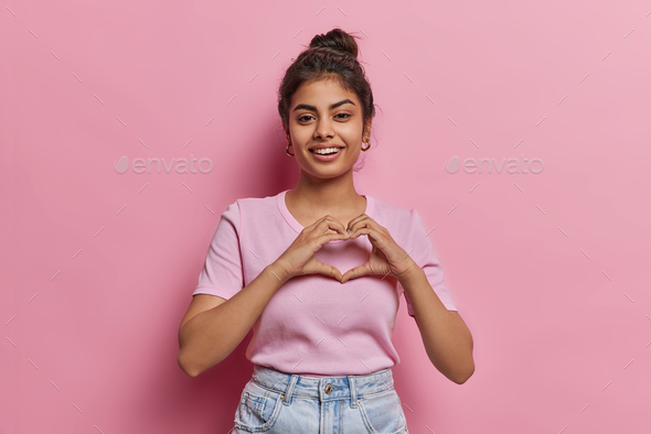 Portrait of happy Indian woman with hair bun shows finger heart gesture ...