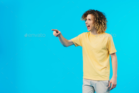 angry curly teenager yelling and pointing with finger isolated on blue ...