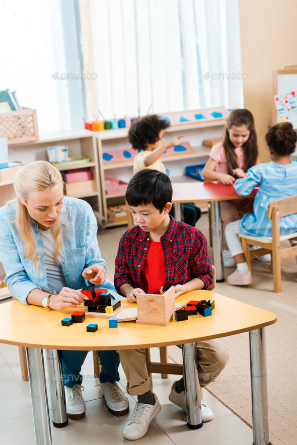 Kid playing building blocks by teacher with children at background in ...
