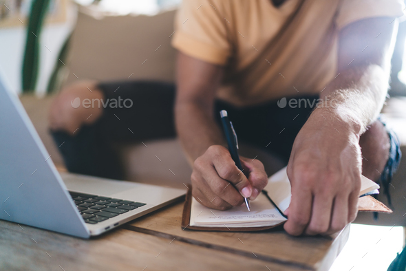Crop man writing in notepad while working on laptop Stock Photo by GaudiLab