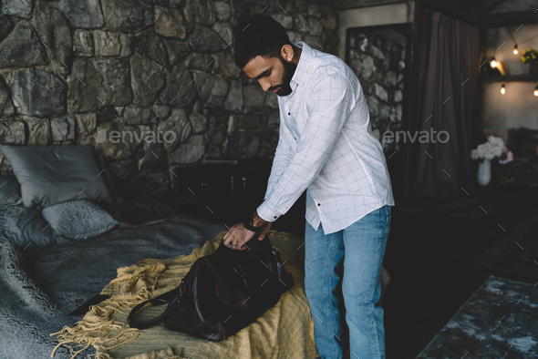 Bearded man unpacking baggage with clothes Stock Photo by GaudiLab