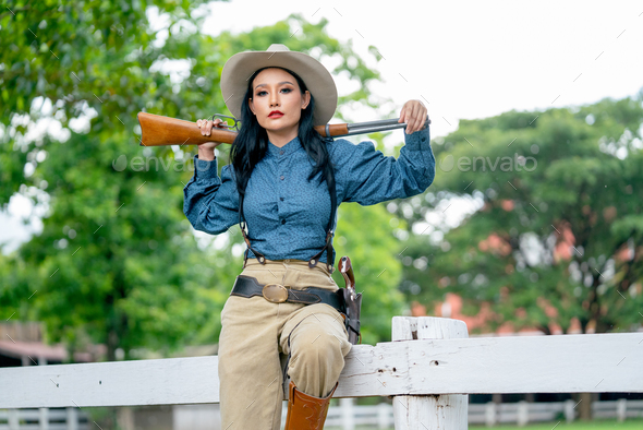 Portrait of pretty Asian woman with cowboy style hold rifle gun on her ...