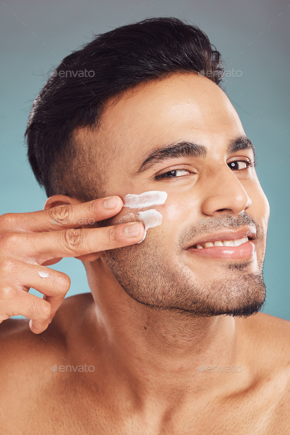 Portrait of one smiling young indian man applying moisturiser lotion to his face while grooming ...