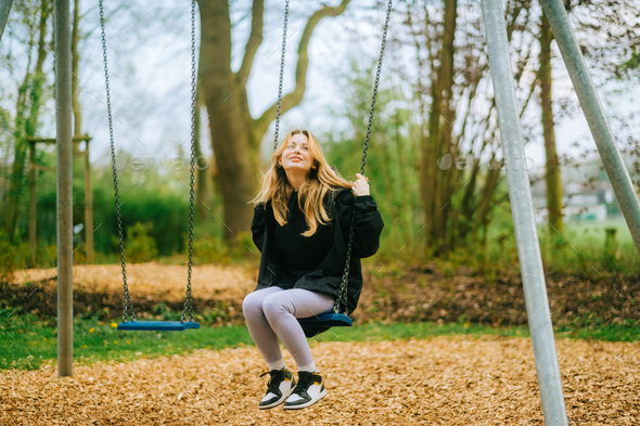 Joyful and youthful woman on a swing at the park. Female in outdoors leisure activity using ...