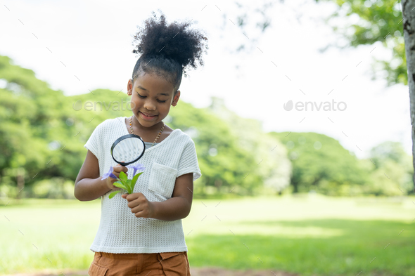 Smiling African American girl holding magnifying glass to explore and look bugs in flower in ...