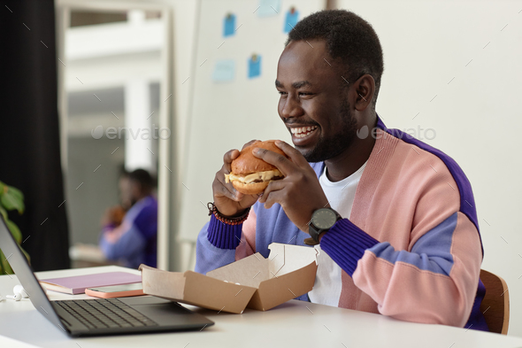 Side view smiling black man eating takeout burger at workplace in ...