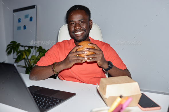 Smiling black man eating burger at workplace indoors Stock Photo by ...